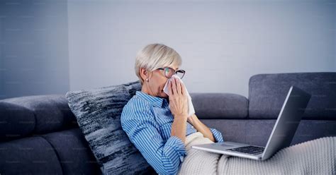 아픈 백인 노인 여성은 담요로 덮인 거실에 앉아 노트북을 무릎에 대고 코를 불고 있습니다 사진 Unsplash의 재채기 이미지