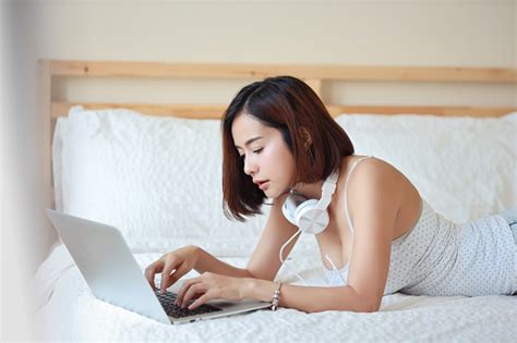 컴퓨터를 사용하고 헤드폰에서 음악을 듣는 동안 침실에서 침대에 누워 흰색 드레스를 입은 아름답고 젊은 성인 아시아 여성의 사진을 클로즈업합니다 귀여운에 대한 스톡 사진 및
