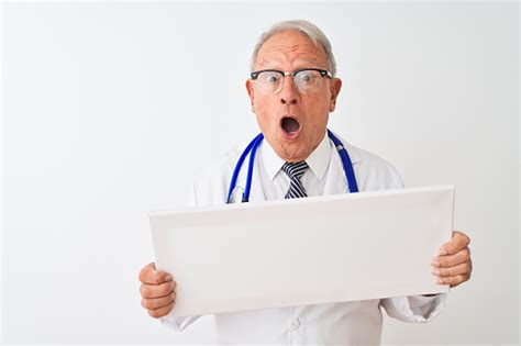 깜짝 얼굴로 충격에 무서워 고립 된 흰색 배경 위에 서있는 배너를 들고 있는 수석 회색 머리 의사 남자 두려움과 두려움 표정으로 흥분 감정에 대한 스톡 사진 및 기타 이미지