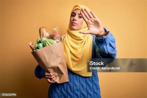 젊은 아름 다운 갈색 머리 아랍 여자 입고 이슬람 히잡 들고 종이 가방 음식 으로 열린 손으로 정지 기호와 심각 하 고 자신감 있는 표정 방어 제스처 식료품에 대한 스톡