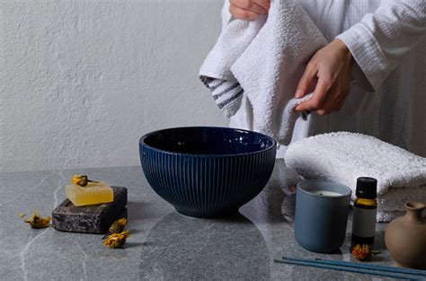 흰색 목욕 가운을 입고 수건 파란색 그릇 천연 바디 케어 제품 회색 표면에 향기 오일을 들고 있는 여성 스파 휴식 건강관리와