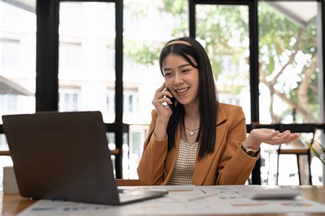 휴대 전화로 컴퓨터 이야기로 일하는 쾌활한 아시아 여성 직원 만족한 비즈니스 여성은 기업 고객과 통화를 하고 재무 보고서 또는 프로젝트 세부 사항을 논의합니다 Office