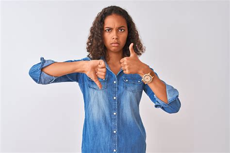 고립 된 흰색 배경 위에 서있는 데님 셔츠를 입은 젊은 브라질 여성은 엄지 손가락을 위아래로 의견 차이와 동의 표현을하고 있습니다 미친 충돌 갈색 머리에 대한 스톡 사진 및