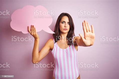 스트라이프 핑크 수영복을 입고 진지하고 자신감있는 표정으로 정지 표지판을하는 열린 손으로 분홍색 배경 위에 말하는 풍선을 들고있는 아름다운 여성 갈색 머리에 대한 스톡 사진