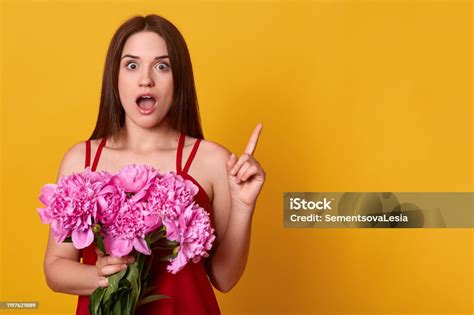 그녀의 손에 장미 빛 모란의 꽃다발과 함께 서 아름다운 소녀 그녀의 검지 손가락이 노란색 배경 위에 고립 된 빈 공간을 가리키는 입을 넓게 열어 유지 세인트 발렌타인 데이