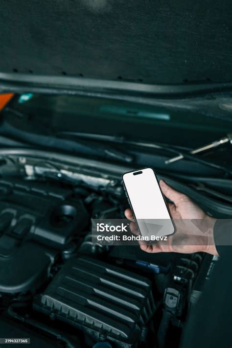 진단 또는 유지 보수를 위해 열린 자동차 엔진 앞에서 빈 흰색 화면이 있는 스마트폰을 들고 있는 정비사 실내 작업장 설정 Brand Name Smart Phone에 대한