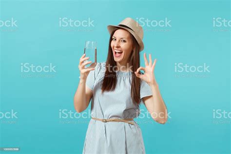 블루 드레스 모자 들고와 유리 파란색 배경에 고립에서 분명 신선한 순수한 물을 마시는 젊은 아름 다운 여자의 초상화 건강 한 생활양식 사람들이 진심으 감정 개념입니다 공간에