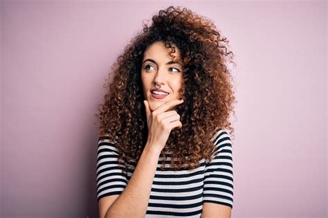 곱슬 머리와 피어싱 캐주얼 스트라이프 티셔츠를 입고 턱에 손을 얹고 질문 펜싱 표현을 생각 하 고 있는 젊은 아름 다운 여자 사려 깊은 얼굴로 미소 의심 개념 T 셔츠에