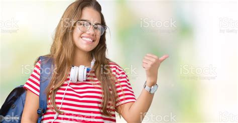 젊은 아름 다운 갈색 머리 학생 여자 보고 하 고 엄지를 가진 측을 가리키는 행복 한 얼굴로 웃 고 고립 된 배경 위에 헤드폰과 배낭을 착용 학생에 대한 스톡 사진 및 기타