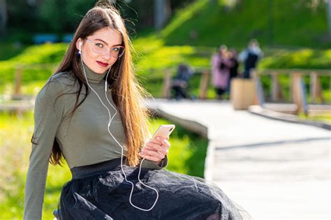 햇볕이 잘 드는 날에 휴대 전화를 사용하여 헤드폰으로 음악을 듣는 야외 공원에서 벤치에 앉아 있는 젊은 미소 짓는 여성 갈색 머리에 대한 스톡 사진 및 기타 이미지 Istock