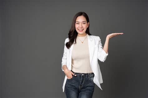 긴 머리를 가진 아시아 여성 임원이 옆으로 서 있다 손을 들어 발표하고 목걸이를 하고 흰 양복과 청바지를 입고 회색 배경 스튜디오에 서서 아름답게 미소를 지었다 25 29