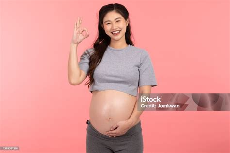 행복한 임산부 서있는 미소는 사랑으로 큰 배를 쓰다듬고 분홍색 배경에 고립 된 Ok를 만들고 젊은 여성의 임신은 미래의 삶 모성 및 임신 개념으로 즐깁니다 임신에 대한 스톡
