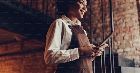 낙관적 인 갈색 머리 숙녀 Witeress는 셀룰러를 손에 들고 사랑스러운 카페 안에 서 있습니다 사진 Unsplash의 사람 이미지