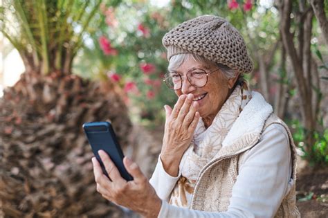야외에 앉아있는 동안 휴대 전화로 화상 채팅에서 웃고있는 모자를 가진 행복한 노인 여성 공원에서 쉬고있는 매력적인 백인 노인 여성 70 79세에 대한 스톡 사진 및 기타