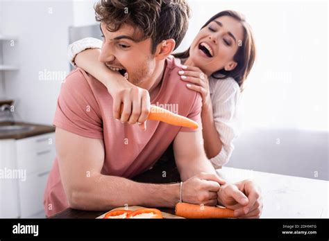 Man Biting Hand Of Cheerful Girlfriend Holding Carrot In Kitchen Stock