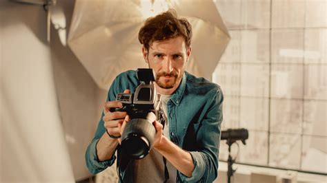 전문 장비가있는 사진 스튜디오에서 배경에서 깜박이는 소프트 박스로 사진을 찍는 최첨단 카메라를 들고있는 유명한 사진 작가의 초상화 Performing Arts Event에