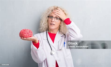 젊은 금발 의사 여자 머리에 손으로 압박 하는 뇌를 들고 그런 지 회색 벽에 충격 수치심과 깜짝 얼굴로 분노와 좌절 두려워 하 고 실수에 대 한 화가 감정에 대한 스톡 사진