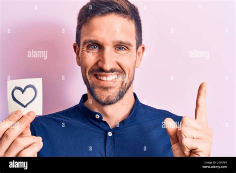 Young Handsome Man Holding Reminder Paper With Heart Shape Over Pink Backgroud Smiling Happy