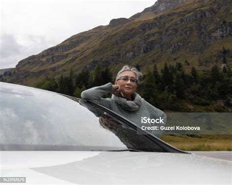 따뜻한 스웨터를 입은 젊은 회색 머리 여성이 고산 계곡의 차 근처에 서 있습니다 60 64세에 대한 스톡 사진 및 기타 이미지