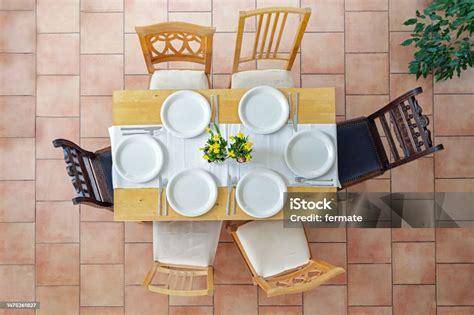 밝은 나무 테이블에 6 개의 흰색 접시 칼 붙이 및 노란색 수선화 꽃과 테라코타 타일 바닥에 6 개의 다른 의자가있는 장소 설정의 상위 뷰 식탁에 대한 스톡 사진 및 기타
