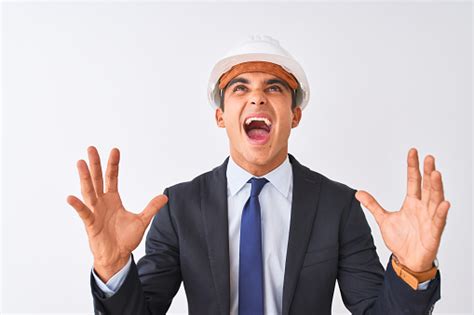 고립된 흰색 배경 위에 양복과 헬멧을 쓰고 있는 젊은 잘생긴 건축가 남자는 공격적인 표정과 팔을 들고 미친 소리와 고함을 질렀다 좌절 개념입니다 감정에 대한 스톡 사진 및