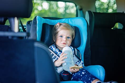 파란 눈을 가진 사랑스러운 여자 아이가 카시트에 앉아 있다 가족 휴가와 조니를 떠나는 유아 교통 체증 중에 행복한 아이를 웃고 병에서 우유를 마시고 비스킷을 먹는다 교통에