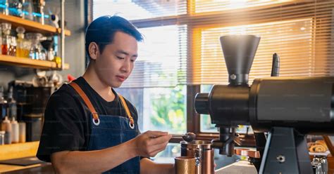 아시아 남자 바리스타가 커피 그라인더 기계를 사용하여 카페의 카운터 바에서 볶은 커피 원두를 갈고 있습니다 남성 커피숍 주인이 고객에게 블랙 커피를 양조하고 있다 중소