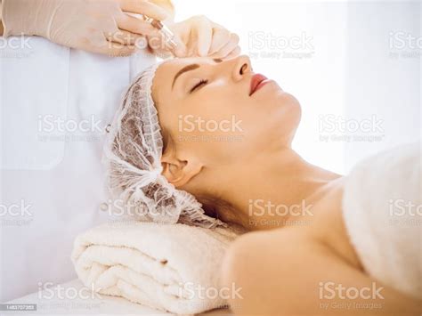 햇볕이 잘 드는 의료 센터에서 눈을 감고 미용 주사를 받는 아름다운 여성 미용사 의사는 주사기로 여성의 얼굴에 미용 시술을 하고