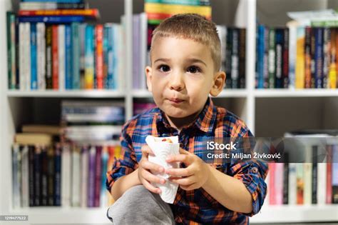 낮이나 아침에 혼자 먹는 샌드위치 음식을 들고 집에서 책장 앞에 서 있거나 서있는 작은 백인 소년 어린 아이의 정면 초상화 셔츠 2 3 살에 대한 스톡 사진 및 기타 이미지