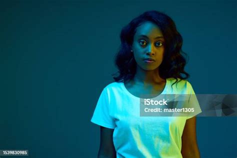 하얀 티셔츠를 입은 젊은 아프리카인 아름다운 여성의 초상화는 네온 불빛의 청록색 파란색 스튜디오 배경에 대해 진지한 얼굴로 카메라를 보고 있다 25 29세에 대한 스톡 사진
