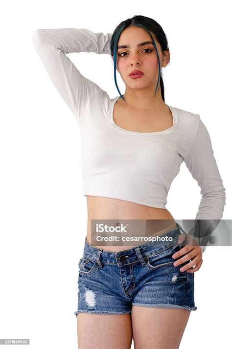 염색 한 푸른 머리와 포니 테일을 한 아름 다운 젊은 여성의 초상화 긴 소매 크롭 탑과 짧은 청바지를 입고 엉덩이에 손을 얹고 흰색 배경에 머리 뒤에 손을 얹고 있습니다