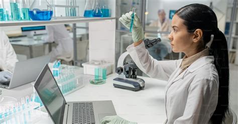 흰 코트를 입은 젊은 진지한 여성 실험실 노동자가 노트북 앞에 직장에 앉아 플라스크에 담긴 파란색 액체를 보고 있다 사진 Unsplash의 실험실 코트 이미지