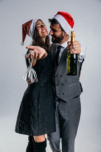 축제 분위기의 스튜디오 배경 속에서 비즈니스 복장을 한 잘 차려입은 커플이 색종이 조각과 샴페인으로 둘러싸인 산타 모자를 쓰고 축하 행사에 기발한 느낌을 더합니다 12월