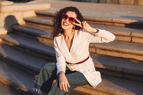 선글라스와 흰색 재킷을 입은 어두운 곱슬 머리를 가진 즐거운 아가씨가 거리의 계단에 앉아 두 손가락 제스처를 보여주면서 카메라를 즐겁게 바라보고 있습니다 20 24세에 대한
