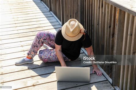 밀짚모자를 쓴 여자가 여름에 나무 다리 바닥에 앉아 노트북으로 작업하고 있는 모습 디지털 노마드 시리즈 It 지원에 대한 스톡 사진 및 기타 이미지 Istock