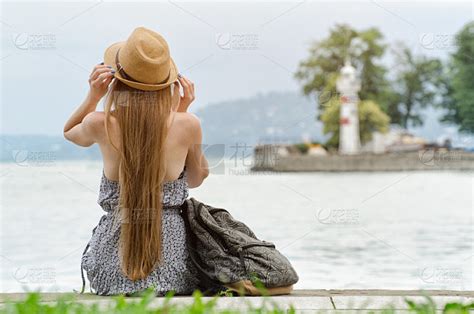 戴着帽子背着背包坐在码头上的女孩背景上是山脉和灯塔从后面看