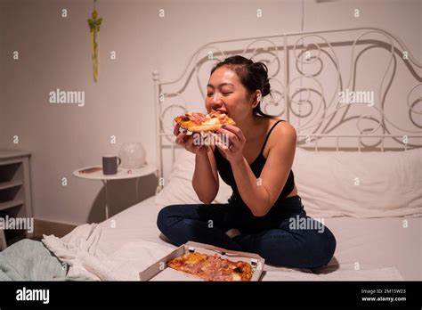 Ethnic Female In Homey Clothes Sitting On Bed With Crossed Legs While Having Delicious Pizza