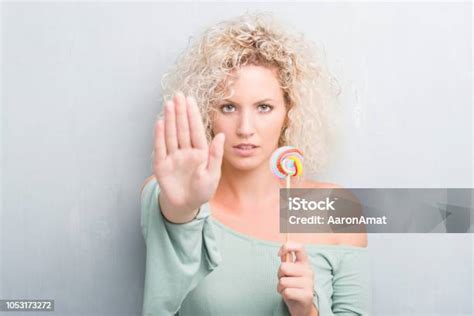 심각 하 고 자신감 표현 방어 제스처 정지 표시를 하 고 오픈 손으로 사탕 막대 사탕을 먹는 그런 지 회색 벽에 젊은 금발 여자 경계 표지에 대한 스톡 사진 및 기타 이미지