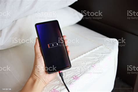 침실에서 배터리 부족 표시기가 있는 충전 중인 스마트폰을 들고 있는 손 Usb 케이블에 대한 스톡 사진 및 기타 이미지