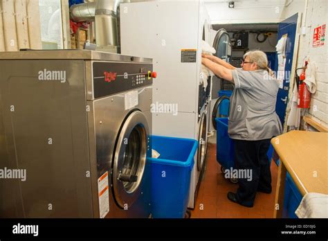 A Woman Working Loading Clothes Into Industrial Size Tumble Dryers In The Laundry Room At A
