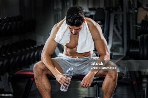 피트 니스 체육관에서 운동 후 벤치 식 수에 앉아 어깨 너머로 흰색 수건으로 피곤 근육 보디 사람 남자 어둠 속에서 휴식 훈련을 복용 하는 벗은 젊은 스포츠 남자 휴식