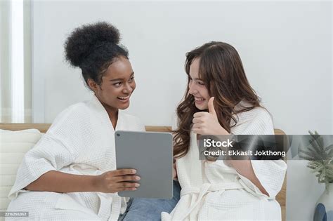 흰 가운을 입은 10대 아프리카계 미국인 및 백인 혼혈 아시아 소녀들이 침대에 앉아 태블릿을 사용하여 기뻐하며 엄지손가락을 치켜세우며 흥분 승인 및 강한 우정을 표현합니다