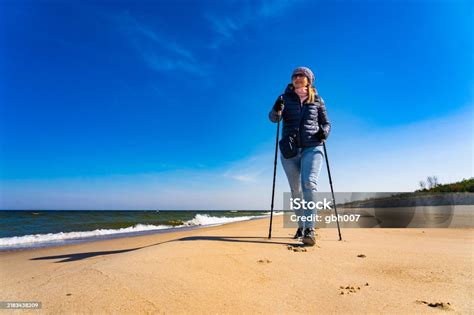 진한 파란색 재킷 청바지 및 겨울 모자를 입고 아름 다운 젊은 여자가 겨울에 화창한 날에 모래 해변을 걷는 북유럽 훈련 앞 View 노르딕 워킹에 대한 스톡 사진 및 기타