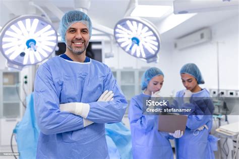 팔을 들고 서 있는 남성 외과 의사의 초상화가 병원의 수술 극장에서 교차했습니다 팀 외과 의사는 수술을 수행하고 중년 의사는 현대 수술실에서 카메라를보고있다 건강관리와