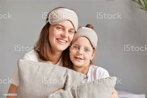 귀여운 딸과 함께 침대에 앉아 있는 쾌활한 어머니는 베개를 들고 미소를 지으며 카메라를 바라보며 자유 시간을 함께 즐기고