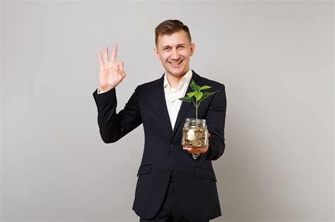 회색 배경에 고립 된 녹색 식물 새싹과 유리 항아리에 더미 금화를 들고 확인 제스처를 보여주는 잘생긴 젊은 비즈니스 남자 업적 경력 자산 사업 개념 복사 공간을 모의 합니다