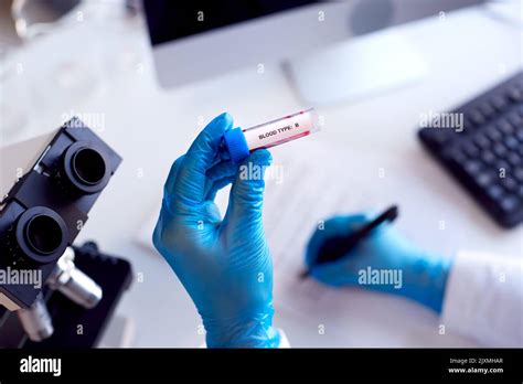 Close Up Of Lab Worker Conducting Research Using Microscope Holding Blood Sample Labelled Type B