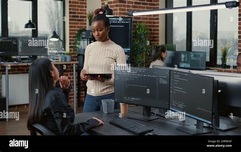 Software Developer Standing Up Holding Digital Tablet Talking With Programer Sitting At Desk