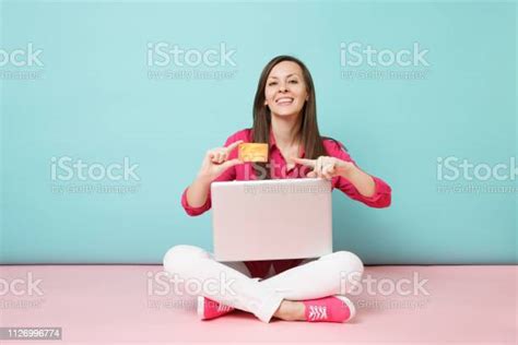 전체 길이 장미 셔츠에 젊은 여자의 초상화 바닥에 앉아 흰 바지 핑크 블루 파스텔 벽 배경 스튜디오에 고립 된 신용 카드 Pc 보유 패션 라이프 스타일 개념입니다 복사