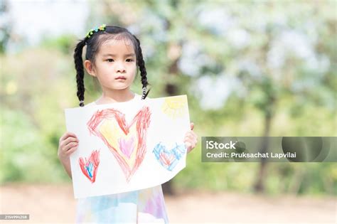 공원에서 종이에 크레용으로 색칠하는 아시아 어린이 소녀 단호한 얼굴을 가진 작고 귀여운 아이가 도화지를 보여주며 재미있는 야외 활동을 합니다 크리에이티브 아이들 교육 아이에